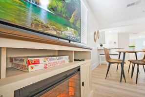 Uma sala de estar moderna com televisão de tela plana sobre a lareira, uma área de refeições com cadeiras de madeira e uma cozinha ao fundo.