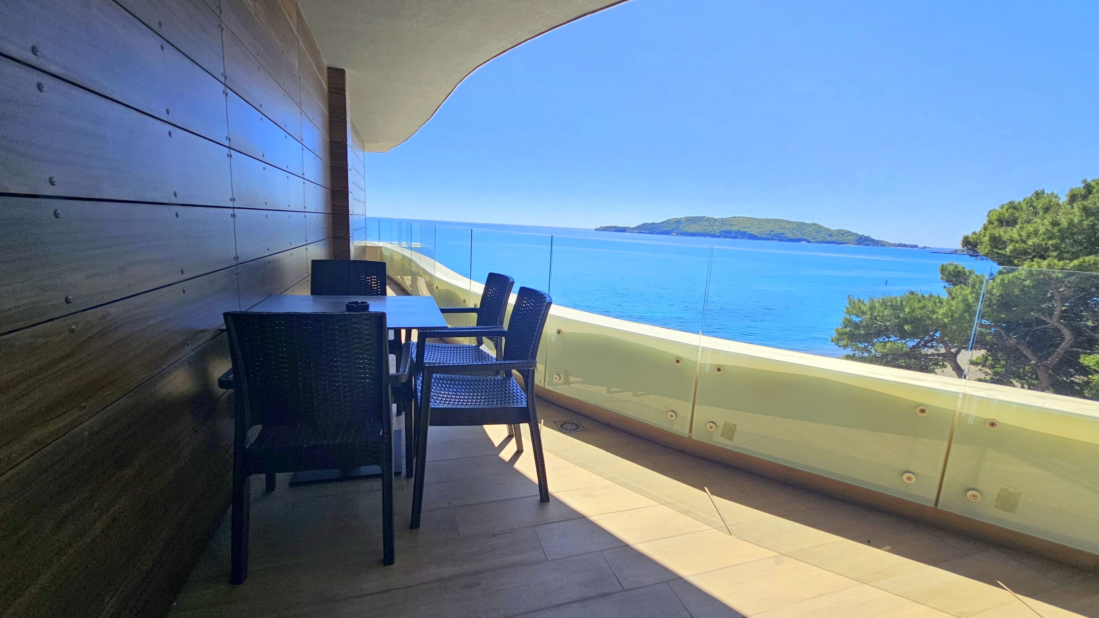 Uma varanda com mesa e cadeiras, com vista para um mar azul límpido e uma ilha distante.