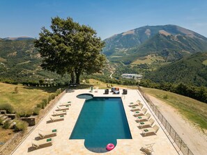 Piscina retangular com espreguiçadeiras e uma árvore em primeiro plano, emoldurada por uma paisagem montanhosa.