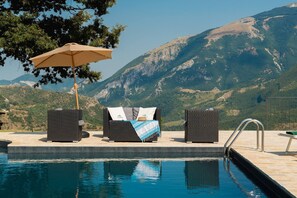 Uma área à beira da piscina com móveis de vime e um guarda-sol, com montanhas ao fundo.