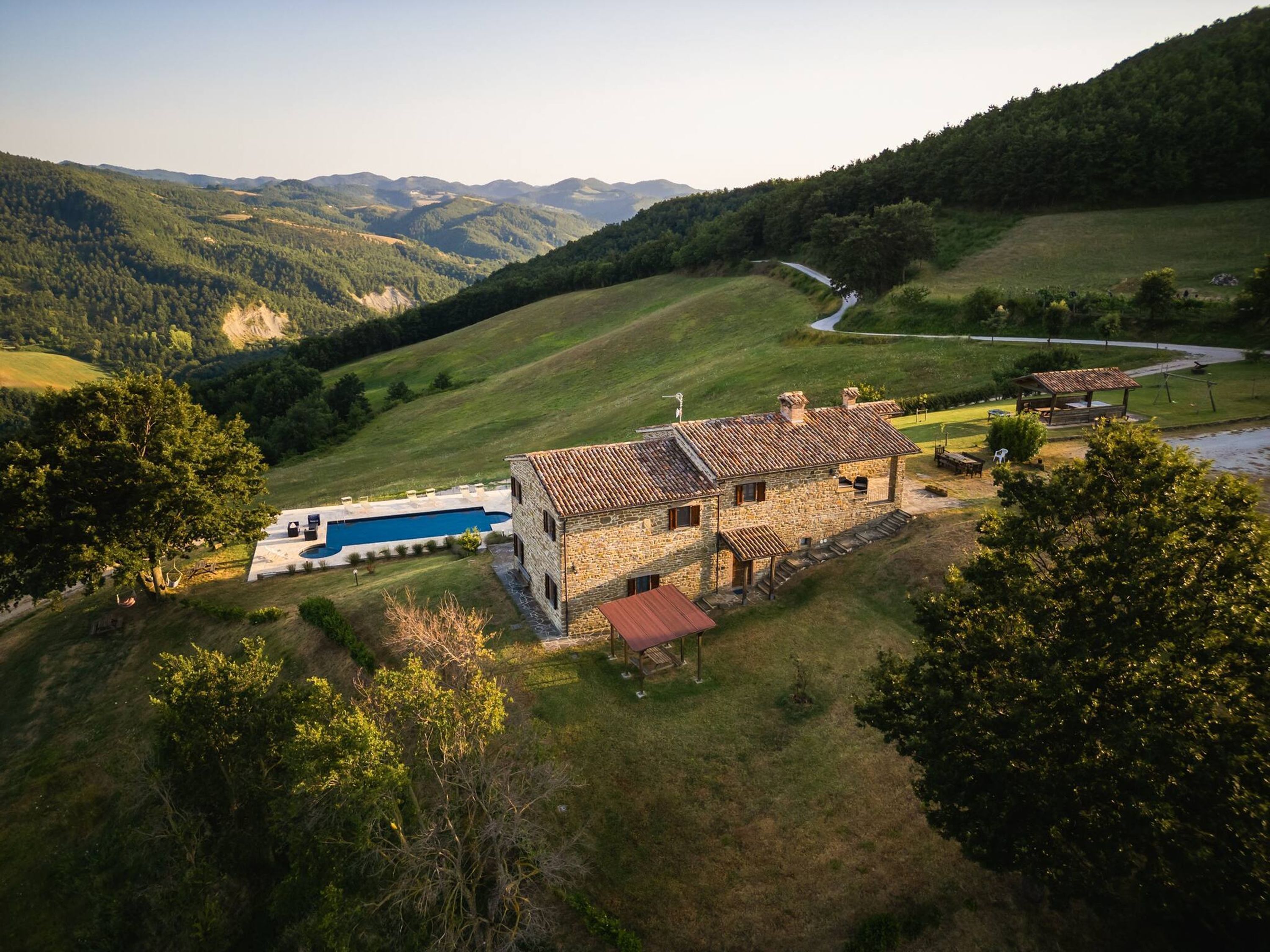 Uma casa de pedra com piscina, rodeada por colinas verdejantes e uma estrada sinuosa.