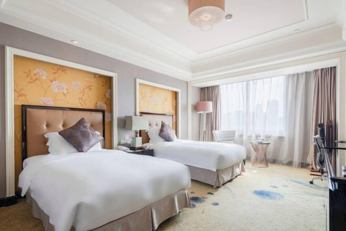 Quarto de hotel com duas camas, uma janela grande com cortinas, uma mesinha de cabeceira e um ventilador de teto.