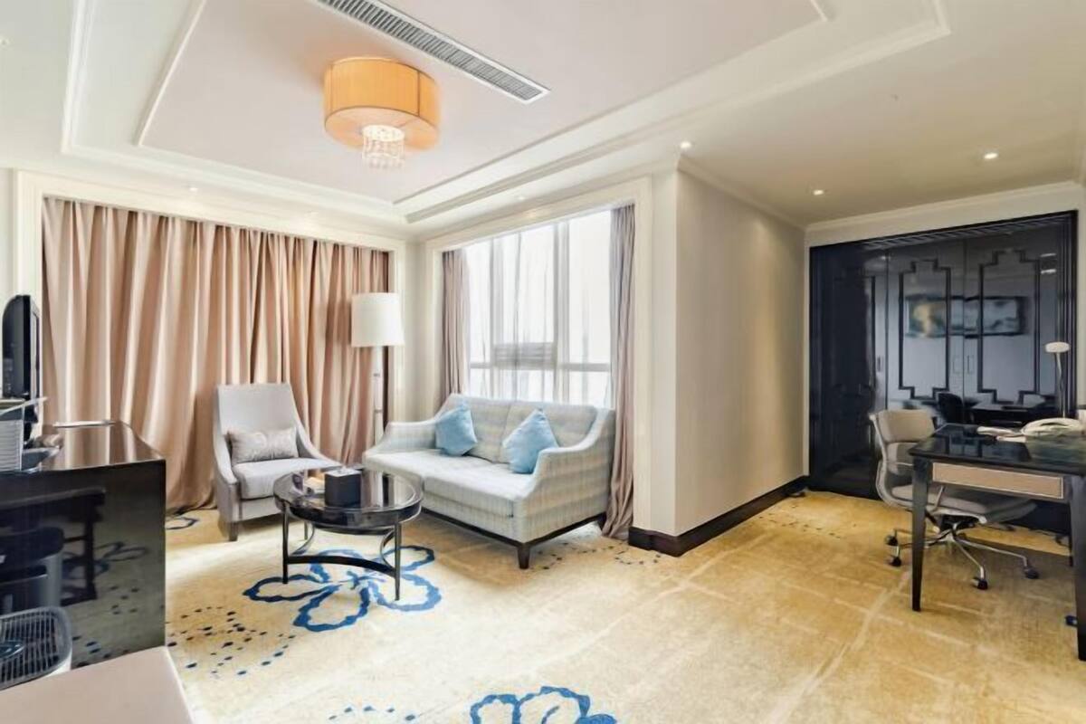 Quarto de hotel com sofá, poltrona, escrivaninha e uma janela grande.