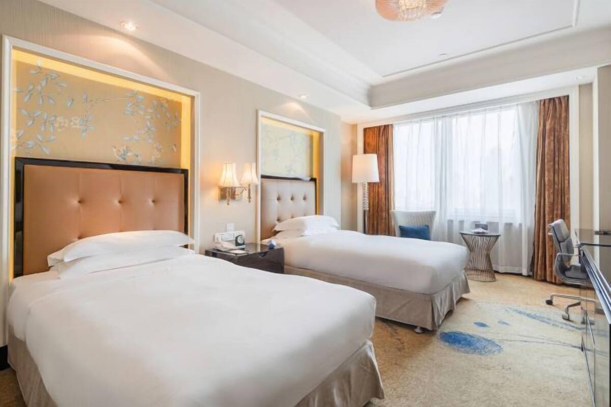 Quarto de hotel com duas camas, uma escrivaninha, uma cadeira e uma mesinha.