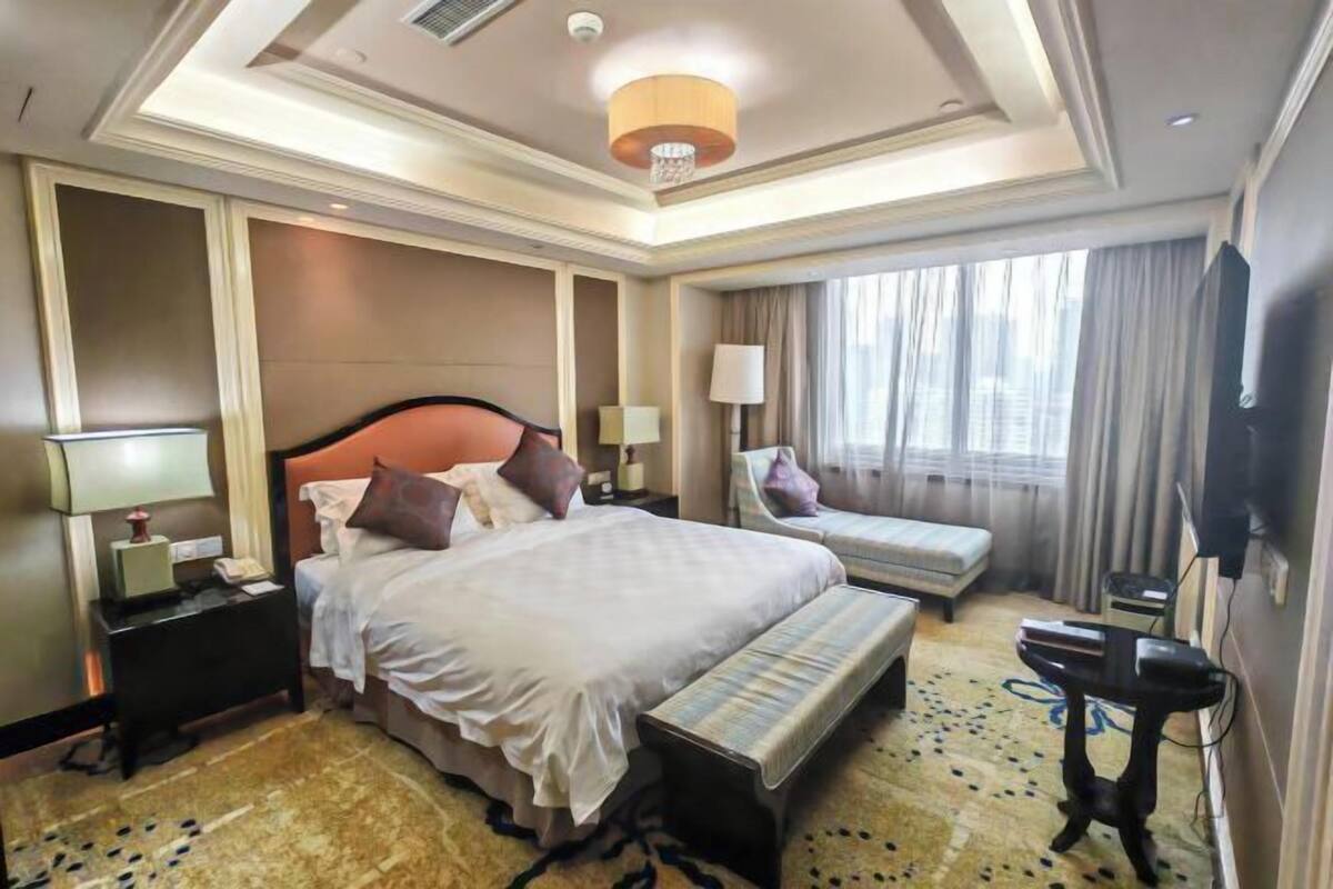 Quarto de hotel com uma cama grande, um banco, uma escrivaninha, uma cadeira, uma TV e uma janela com cortinas.