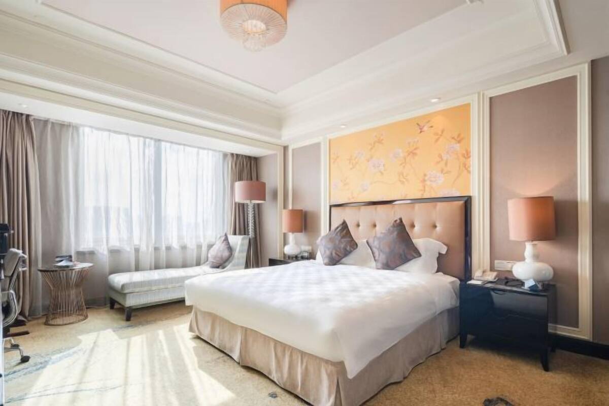 Quarto de hotel com cama grande, mesas de cabeceira, escrivaninha e janela com cortinas.