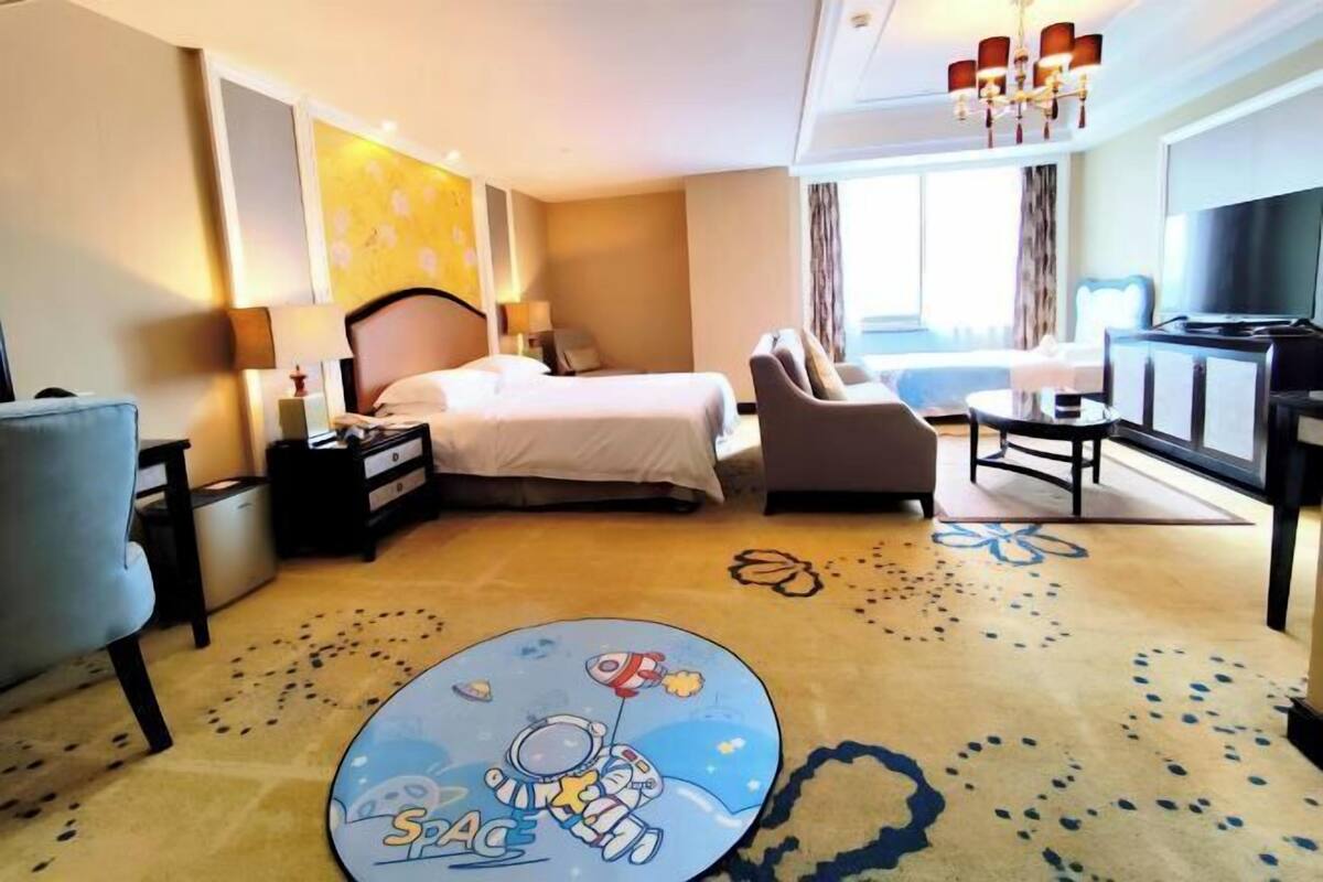 Quarto de hotel com cama, escrivaninha, cadeira, televisão e tapete com tema espacial.