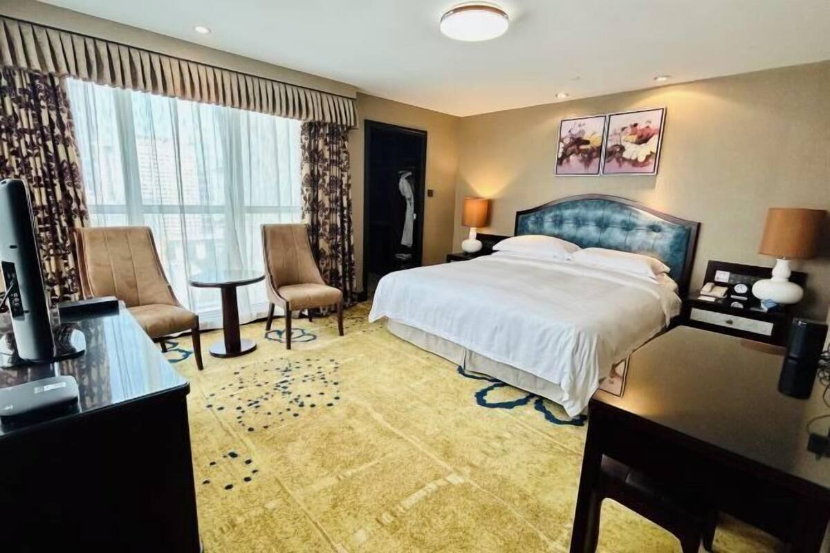 Quarto de hotel com uma cama, duas cadeiras, uma mesinha, televisão e obras de arte na parede.