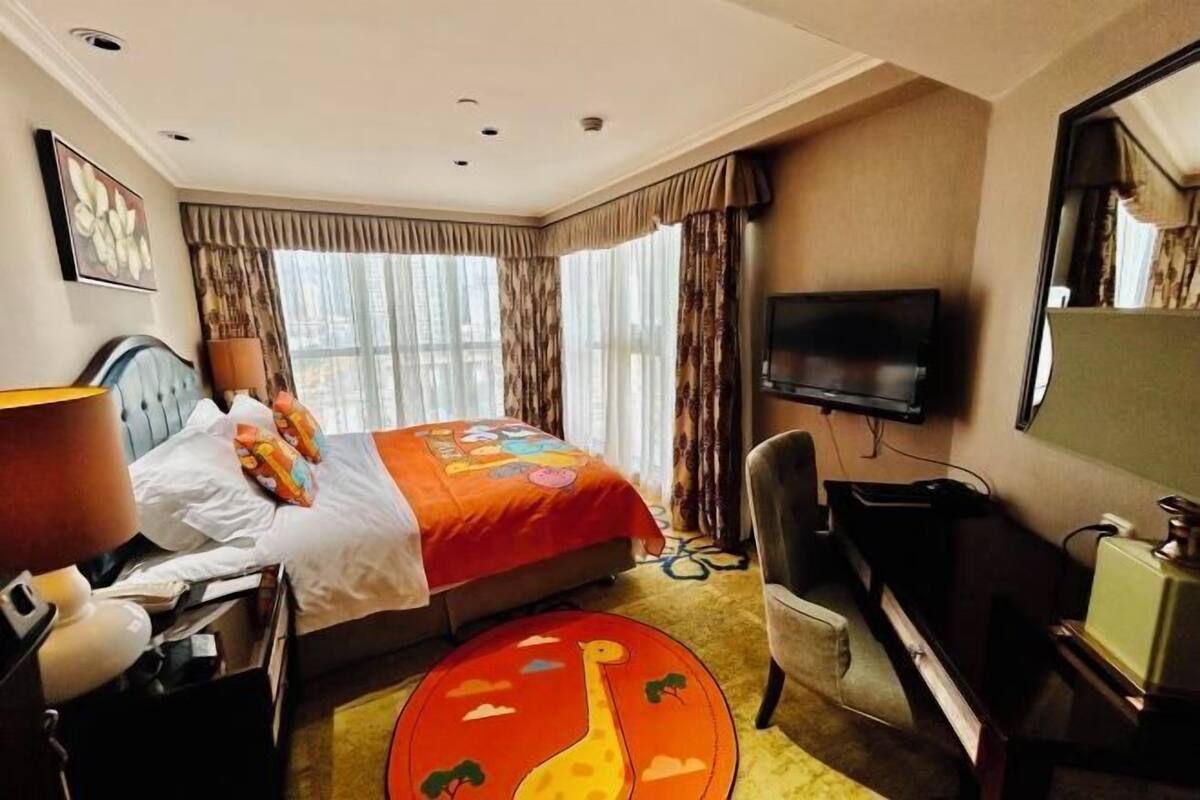 Quarto de hotel com cama, escrivaninha, cadeira, televisão e janela com cortinas.