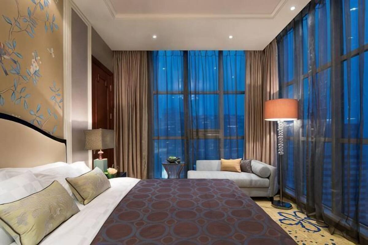 Quarto de hotel com uma cama grande, um sofá, uma mesinha de cabeceira, uma luminária e vista da cidade por grandes janelas.