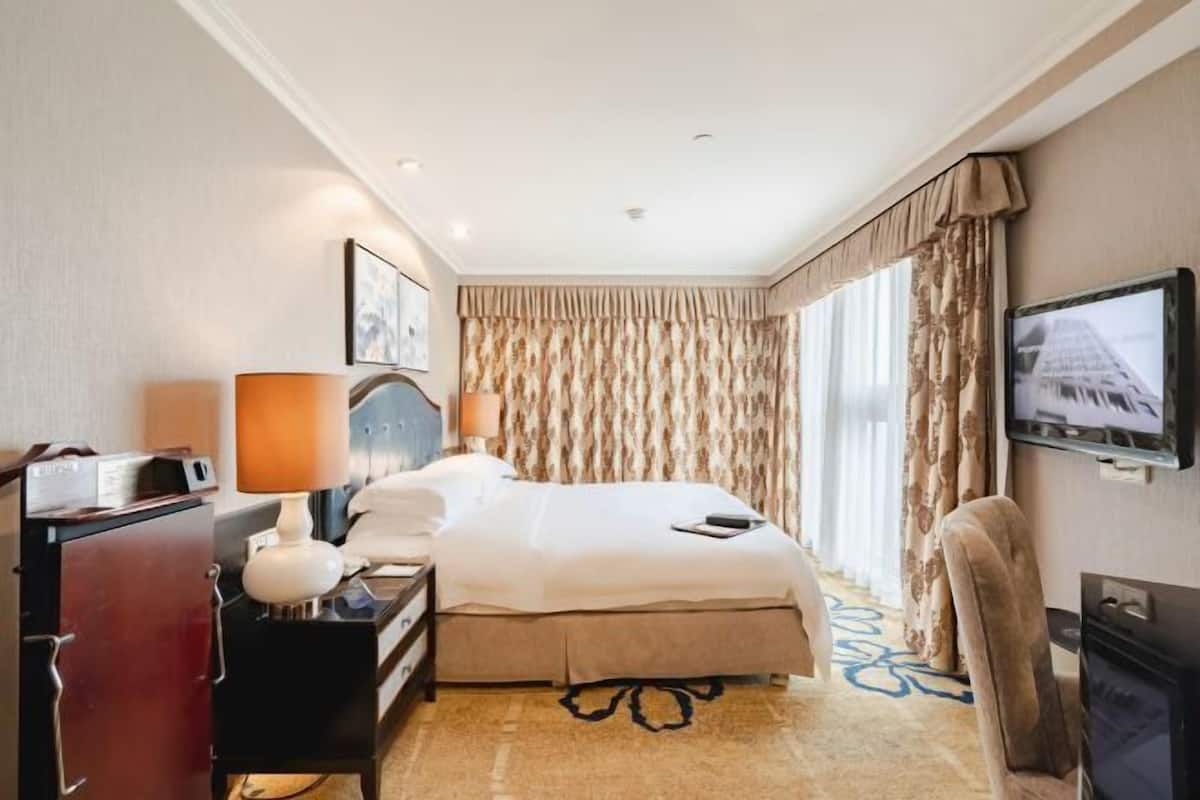 Quarto de hotel com cama, mesa de cabeceira, luminária, televisão e cortinas.