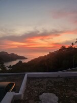 Vista do pôr do sol a partir de uma varanda com vista para a costa, palmeiras e uma ilha distante.