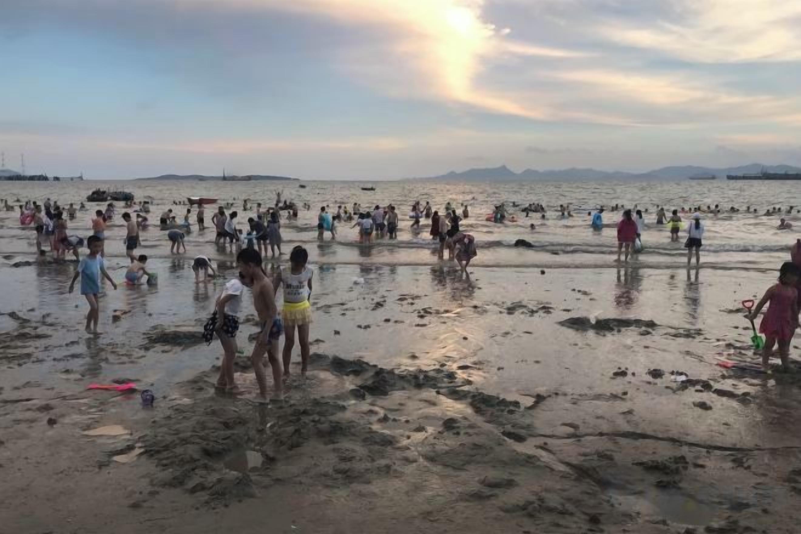 Praia lotada com pessoas na água rasa, barcos ao longe e um pôr do sol ao fundo.