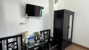 Quarto pequeno e limpo, com televisão de tela plana na parede, um armário preto, uma mesinha com abajur e uma lixeira.