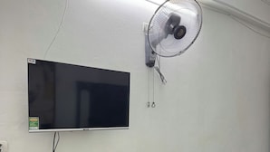 Quarto com televisão de tela plana na parede, um ventilador, uma mesa com luminária, chaleira e um quadro de cardápio.