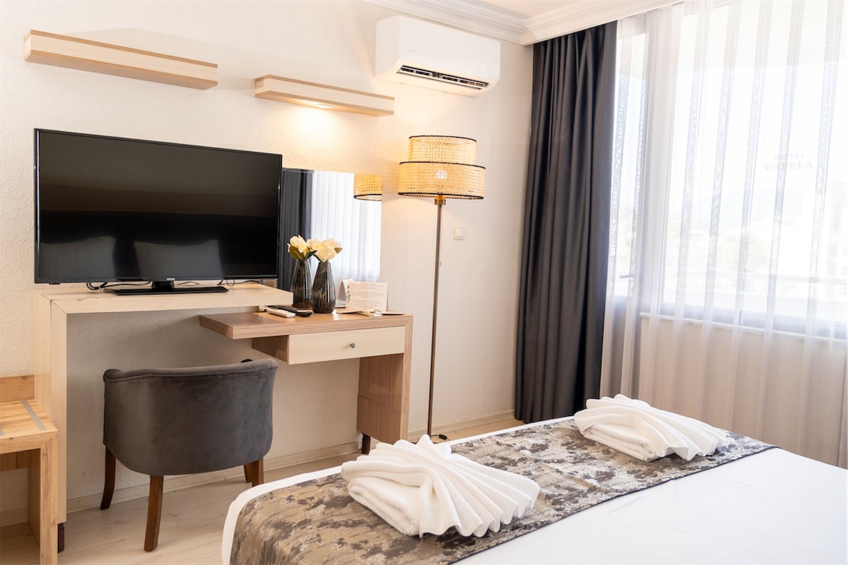 Quarto de hotel com cama, escrivaninha, televisão de tela plana, cadeira, abajur e janela com cortinas.