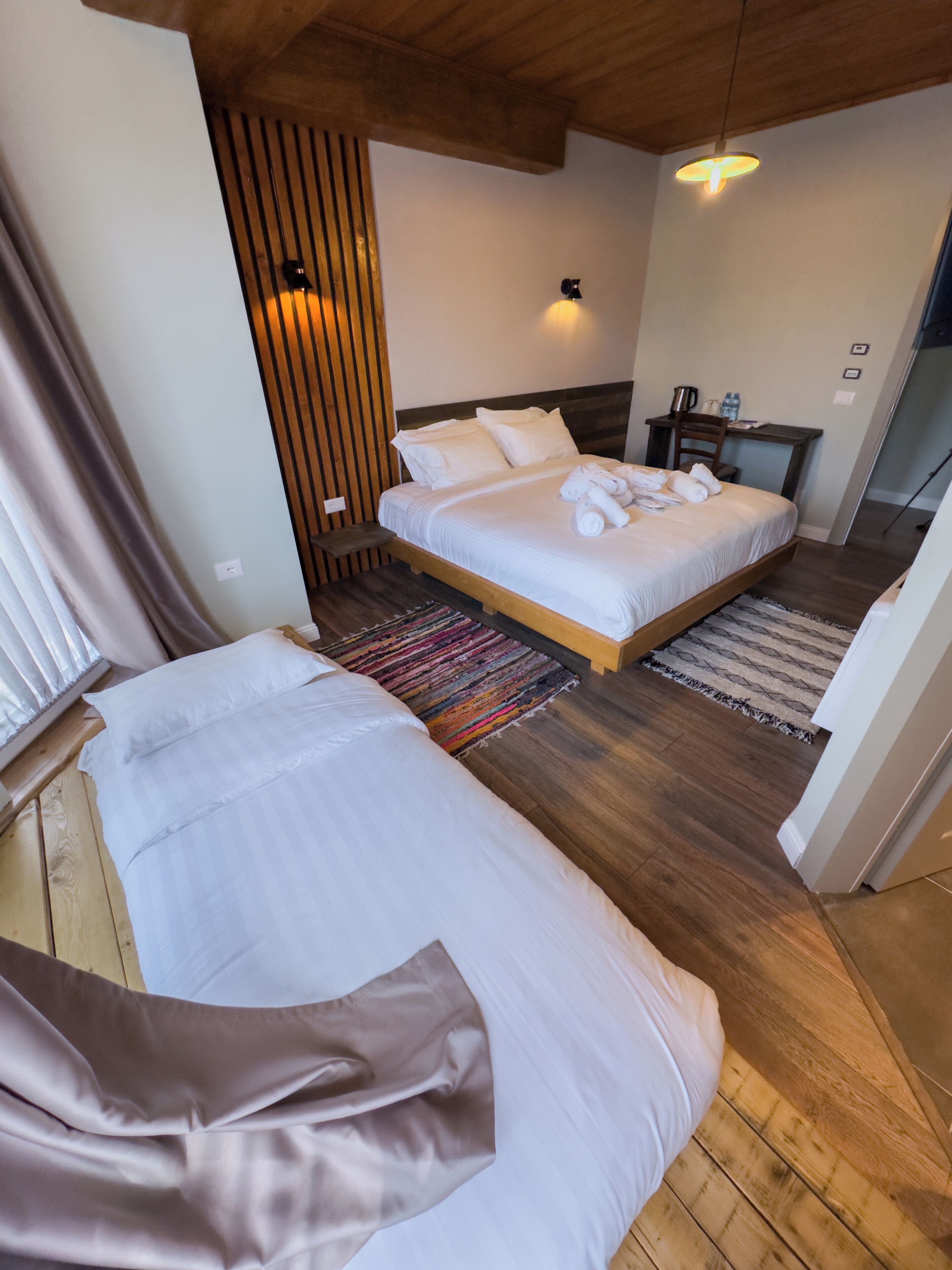 Quarto de hotel com duas camas, teto de madeira, escrivaninha com cadeira e uma pequena mesa com chaleira e garrafas de água.