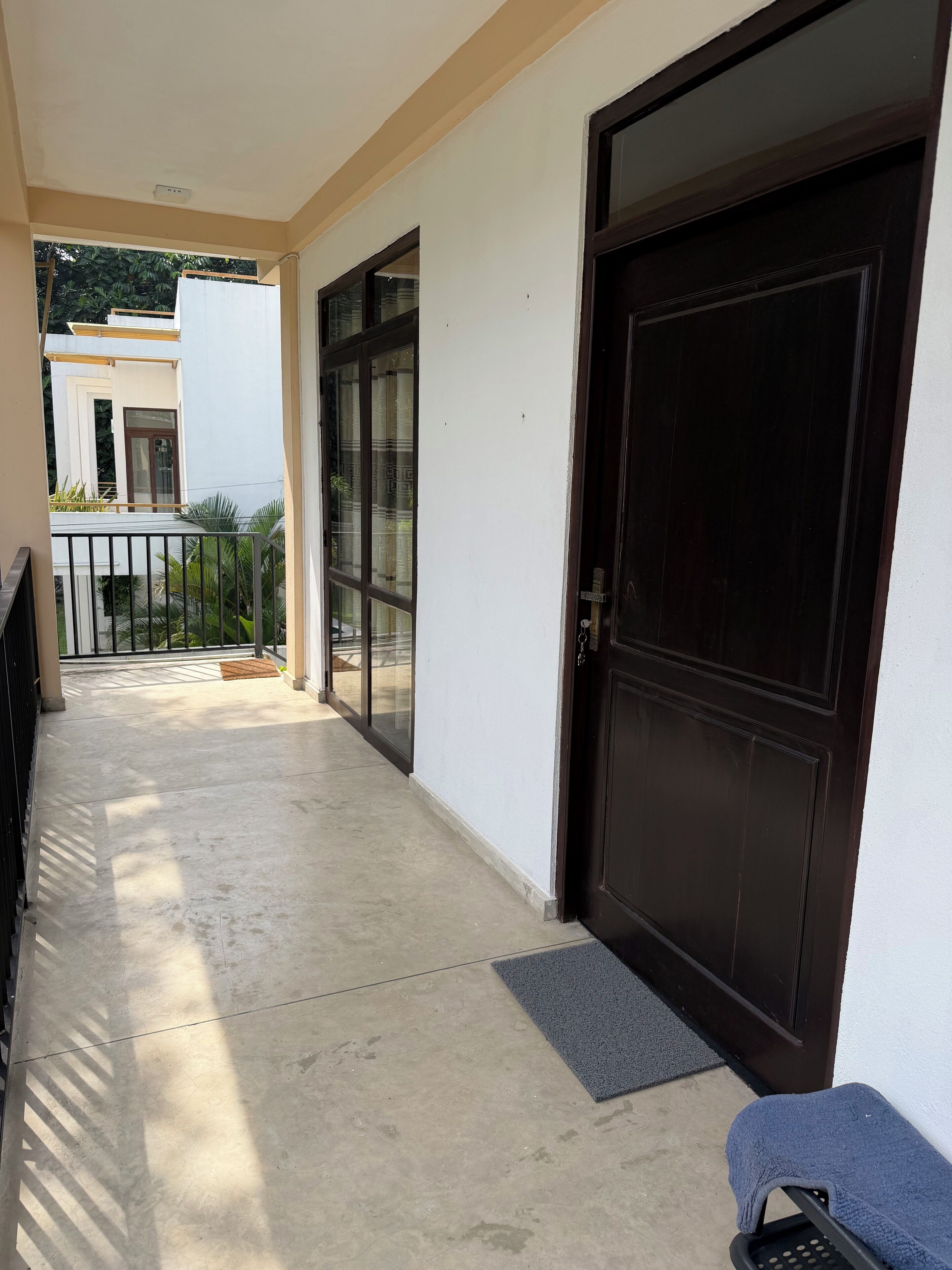 Varanda coberta com piso de concreto, porta de madeira escura e vista para um edifício com portas de vidro.