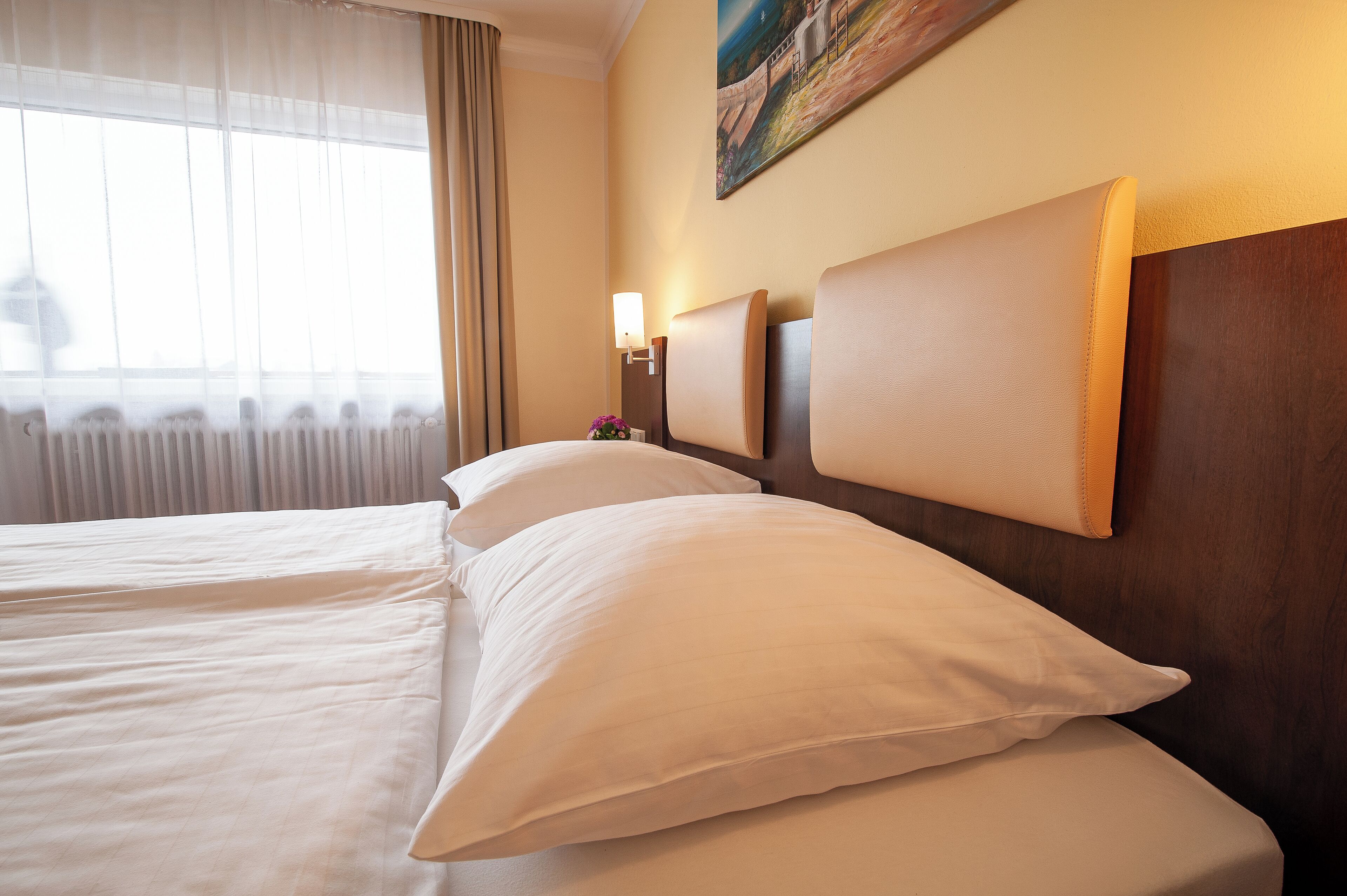 Quarto de hotel com duas camas, uma mesa de cabeceira com abajur, um quadro na parede e uma janela com cortinas.