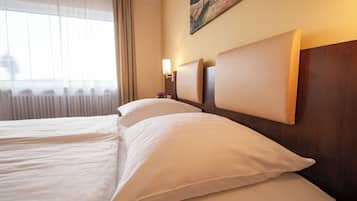 Quarto de hotel com duas camas, uma mesa de cabeceira com abajur, um quadro na parede e uma janela com cortinas.