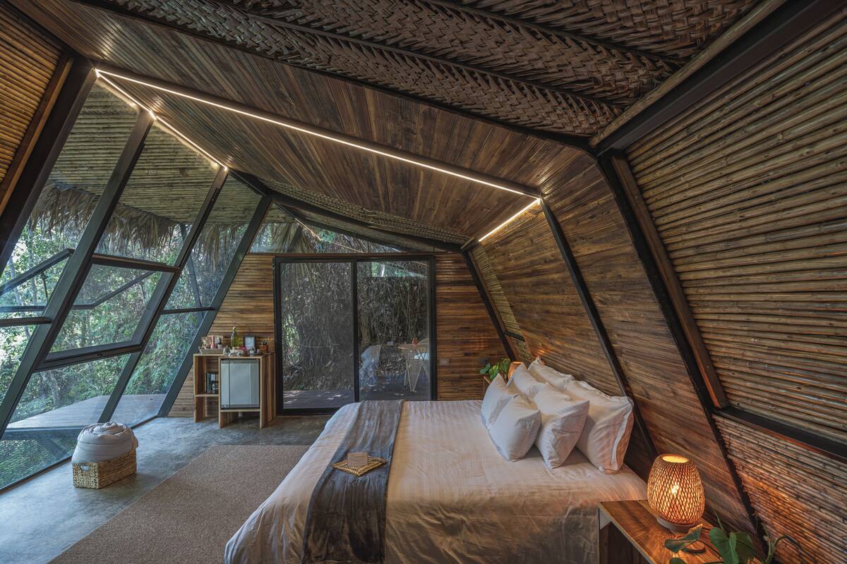 Quarto com uma cama grande, mesa de cabeceira com abajur e vista para o exterior pelas grandes janelas.