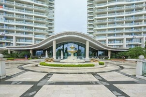 Entrada de hotel com uma fonte, rodeada por área ajardinada e vários edifícios altos ao fundo.