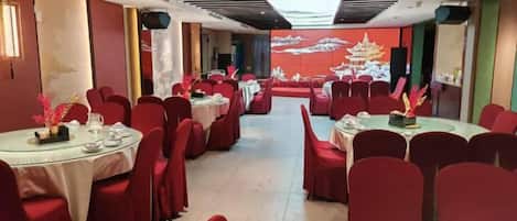 Salão de festas com mesas redondas e cadeiras vermelhas, palco com um cenário tradicional e uma tela exibindo uma cena festiva.