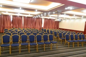 Uma sala de conferências com fileiras de cadeiras, uma tela de projetor e cortinas.