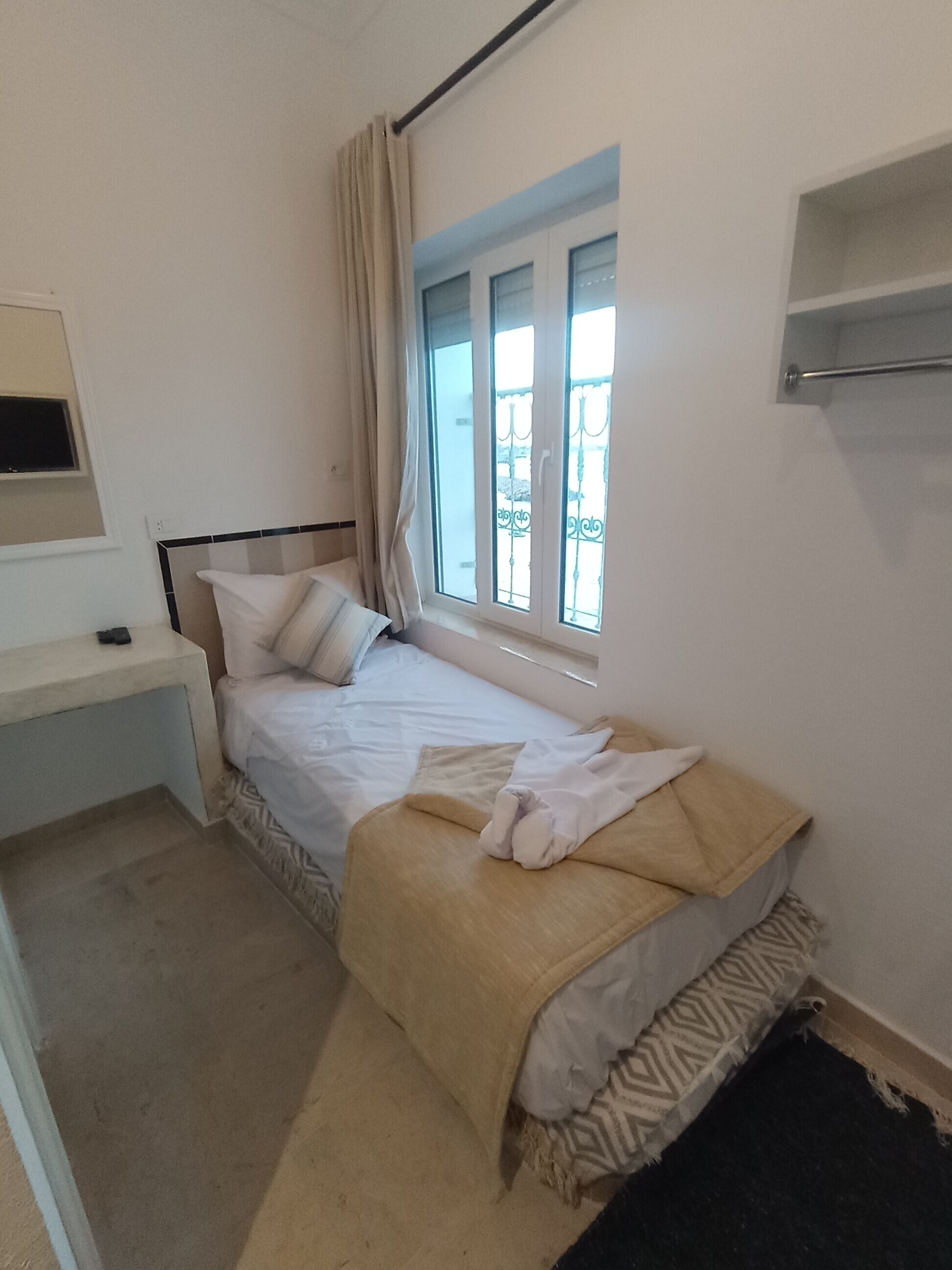 Quarto pequeno e limpo com uma cama de solteiro, mesa de cabeceira, janela com cortinas e prateleira fixada na parede.