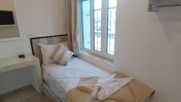 Quarto pequeno e limpo com uma cama de solteiro, mesa de cabeceira, janela com cortinas e prateleira fixada na parede.