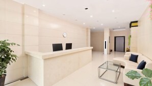 Uma área de recepção com balcão, duas cadeiras, relógio, planta e um sofá com mesa de centro de vidro.
