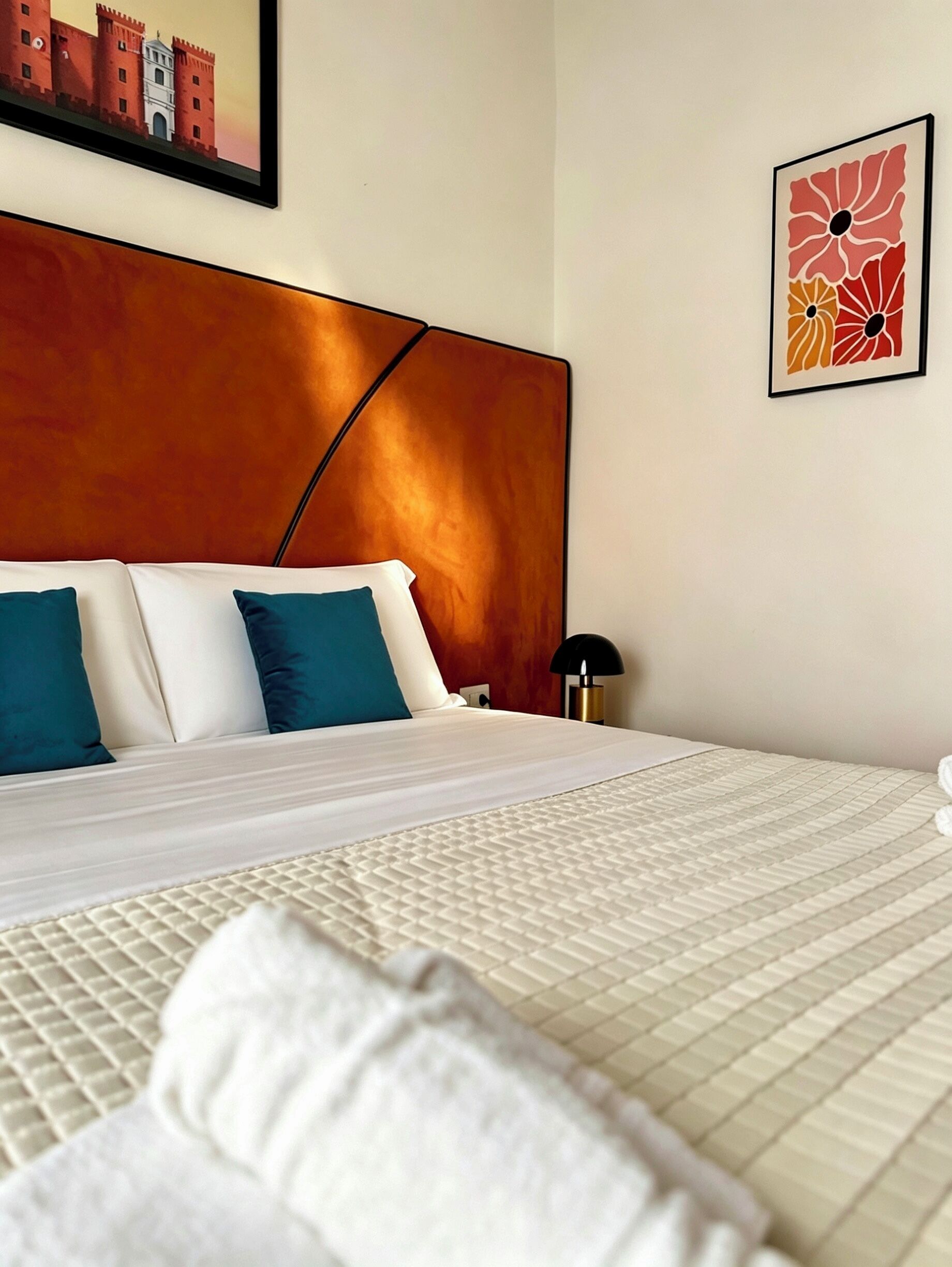 Quarto de hotel com uma cama grande, cabeceira, mesa de cabeceira com abajur e obras de arte nas paredes.