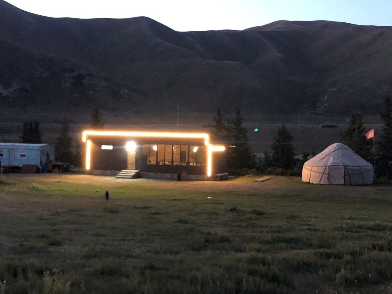 Um edifício moderno com fachada de vidro e entrada bem iluminada, cercado por um campo gramado e uma yurt numa área montanhosa.