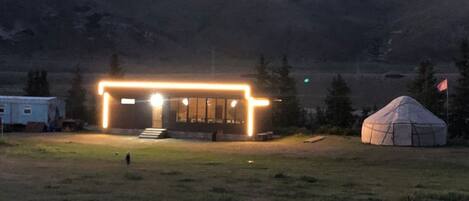 Um edifício moderno com fachada de vidro e entrada bem iluminada, cercado por um campo gramado e uma yurt numa área montanhosa.