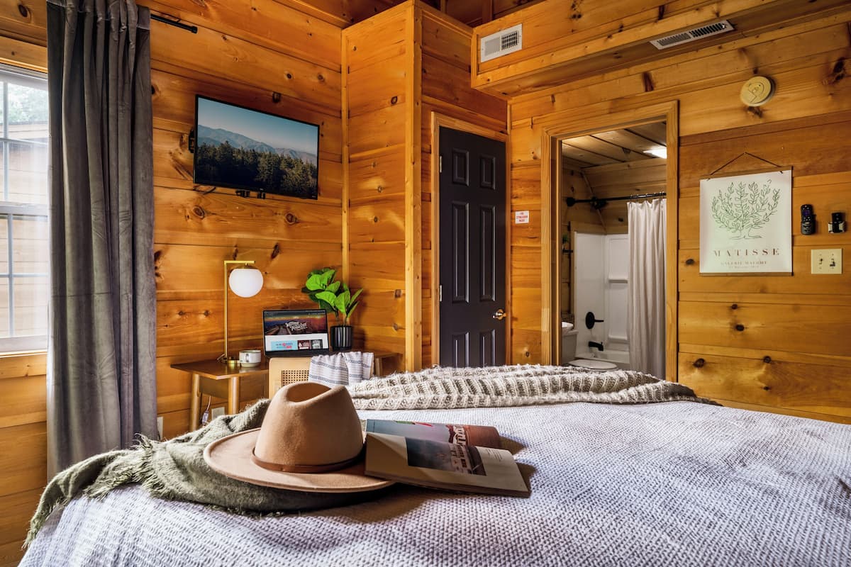 Quarto acolhedor em cabana de madeira, com cama, televisão, uma pequena mesa com uma planta e uma porta que dá para outro quarto.