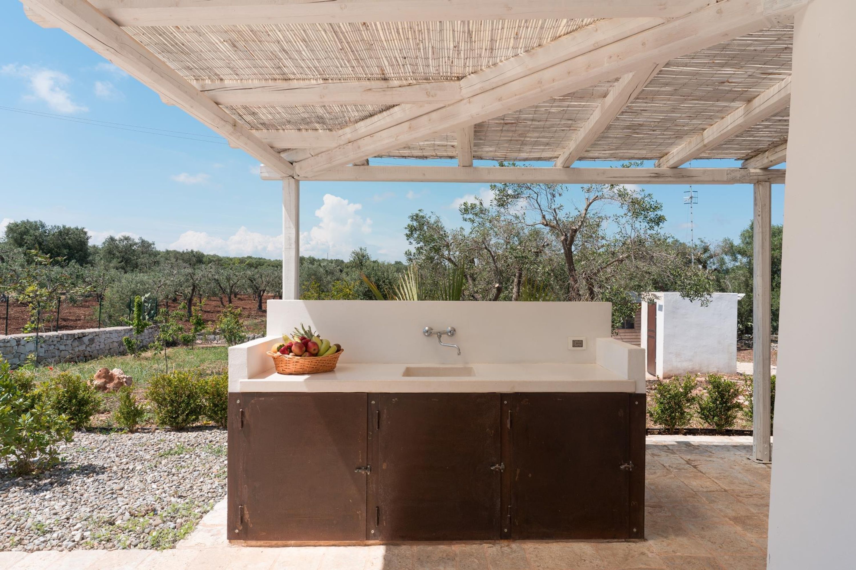 Cozinha ao ar livre com pia e uma cesta de frutas, cercada por um alpendre de pedra e vista para o campo.