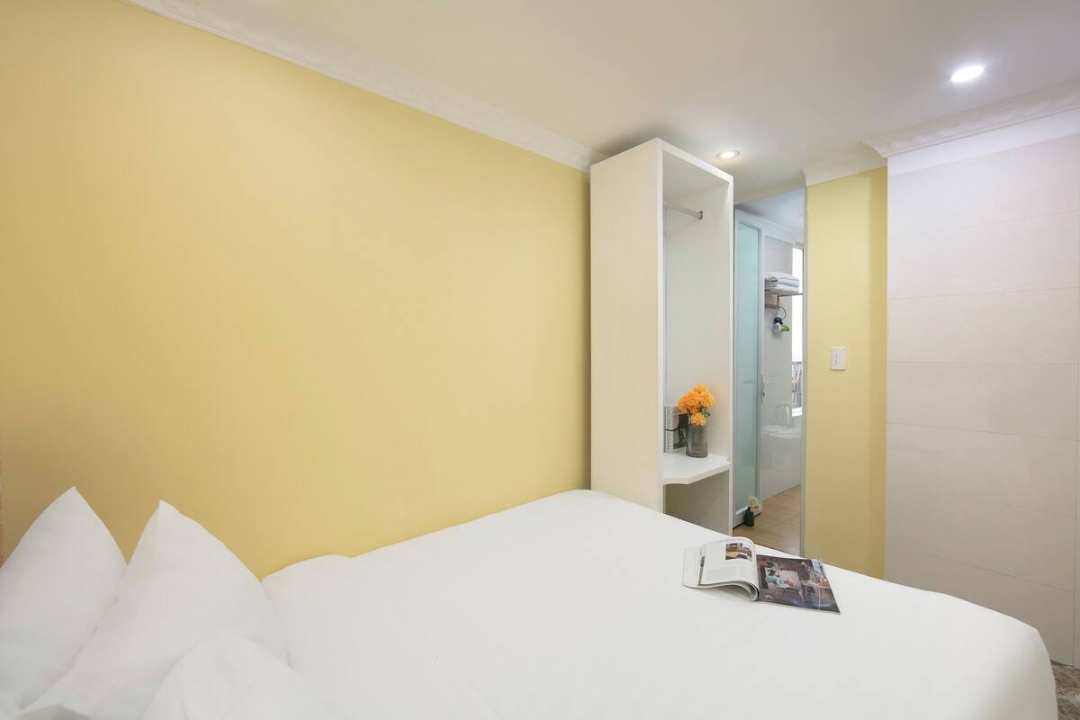 Quarto de hotel com uma cama, mesa de cabeceira branca e uma pequena prateleira com um vaso de flores.