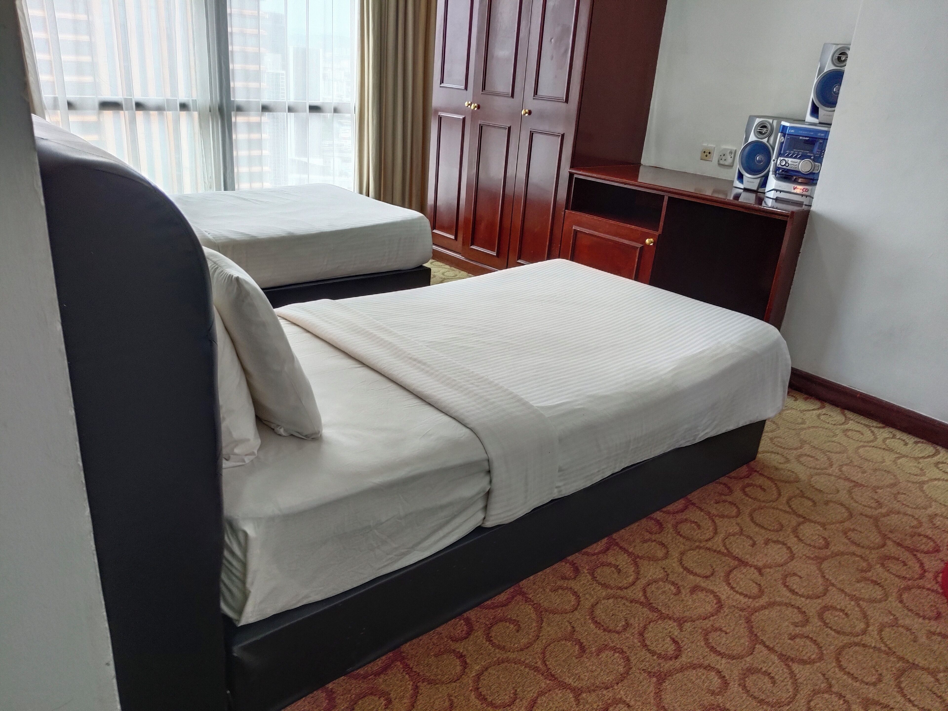Quarto de hotel com duas camas, uma mesa de trabalho com caixas de som e vista da cidade por grandes janelas.