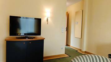 Quarto de hotel com televisão de tela plana sobre um armário de madeira, luminária fixada na parede, porta com espelho e cadeira listrada.