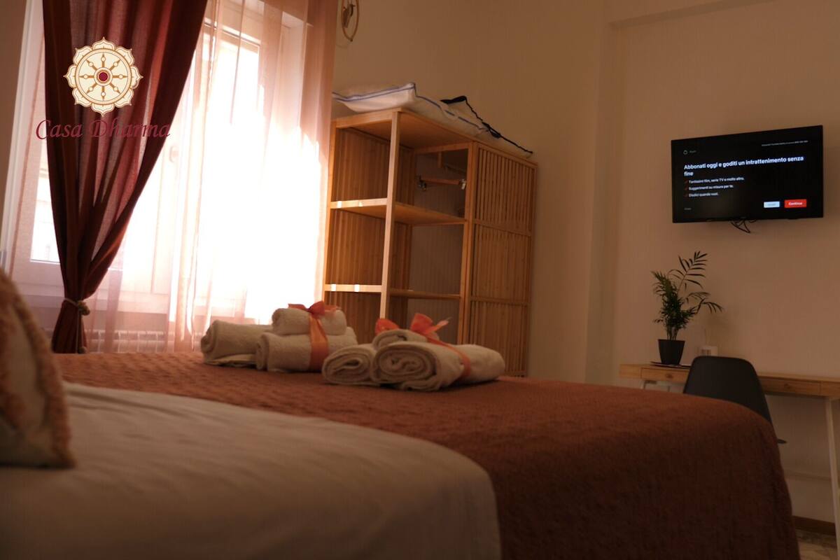 Quarto de hotel com uma cama, um guarda-roupa de madeira, uma mesa com uma planta e uma televisão na parede.
