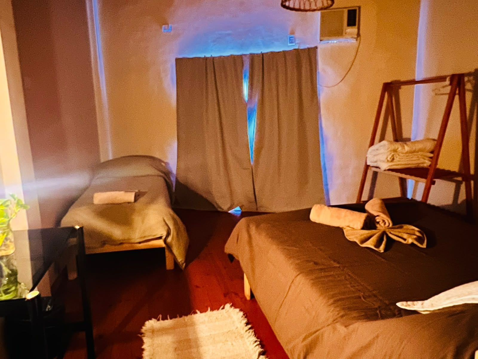 Quarto de hotel com duas camas, uma mesa pequena, uma cadeira, prateleira com toalhas dobradas e uma janela com cortinas.