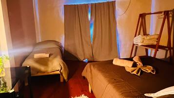 Quarto de hotel com duas camas, uma mesa pequena, uma cadeira, prateleira com toalhas dobradas e uma janela com cortinas.
