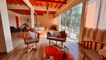 Ampla sala de estar com piso de madeira, teto alto com vigas aparentes e grandes janelas com vista para o exterior.