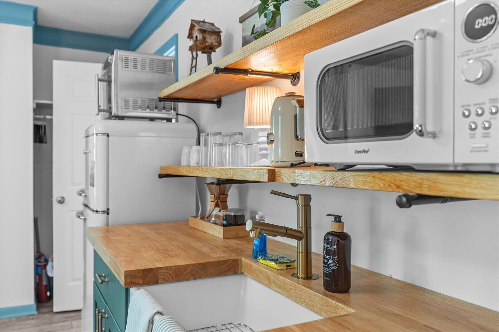 Uma cozinha compacta com geladeira branca, cafeteira, micro-ondas e uma prateleira de madeira com diversos objetos.