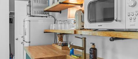 Uma cozinha compacta com geladeira branca, cafeteira, micro-ondas e uma prateleira de madeira com diversos objetos.