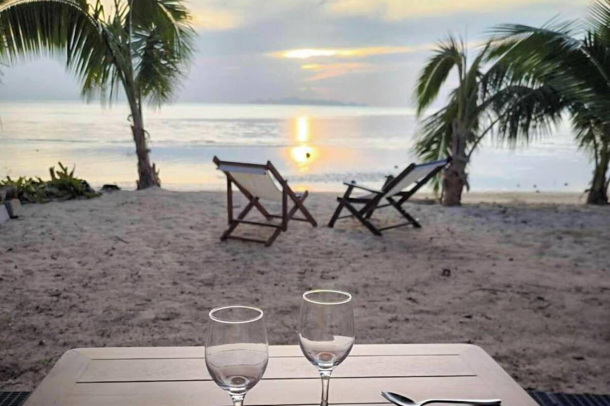 Mesa de madeira posta com pratos, copos e talheres numa praia com palmeiras e vista para o pôr do sol.