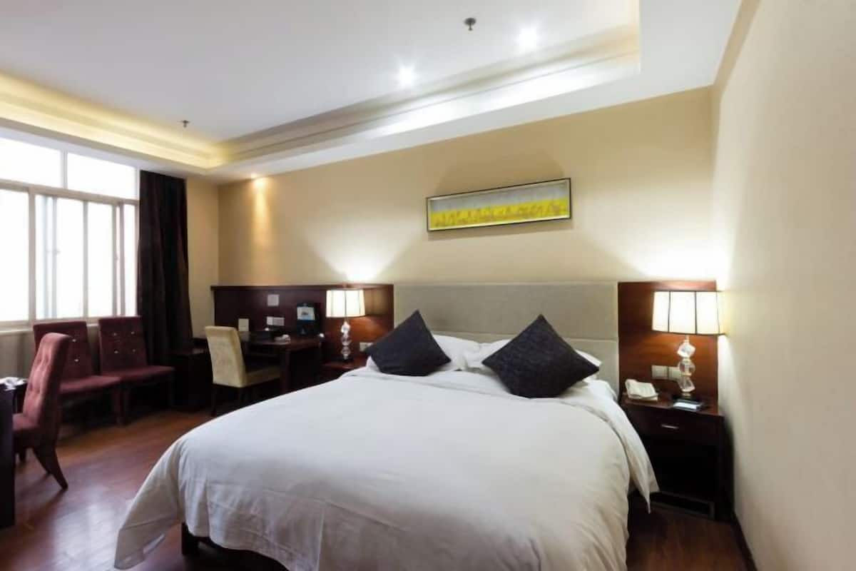 Quarto de hotel com uma cama grande, duas mesas de cabeceira com luminárias, uma escrivaninha com cadeira e uma janela com cortinas.