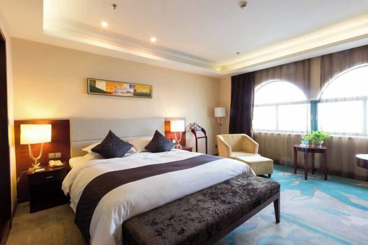Quarto de hotel com uma cama grande, mesas de cabeceira, uma cadeira, um banco, uma escrivaninha e uma janela com cortinas.