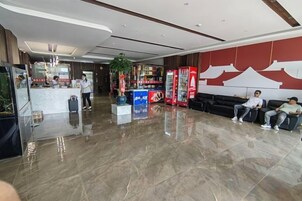 Um saguão de hotel com balcão de recepção, área de estar, máquinas de venda automática e uma grande parede vermelha.