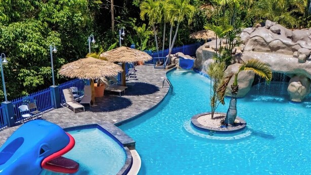 Piscina com escorregador azul, cadeiras de descanso e área de estar coberta por um telhado de palha.